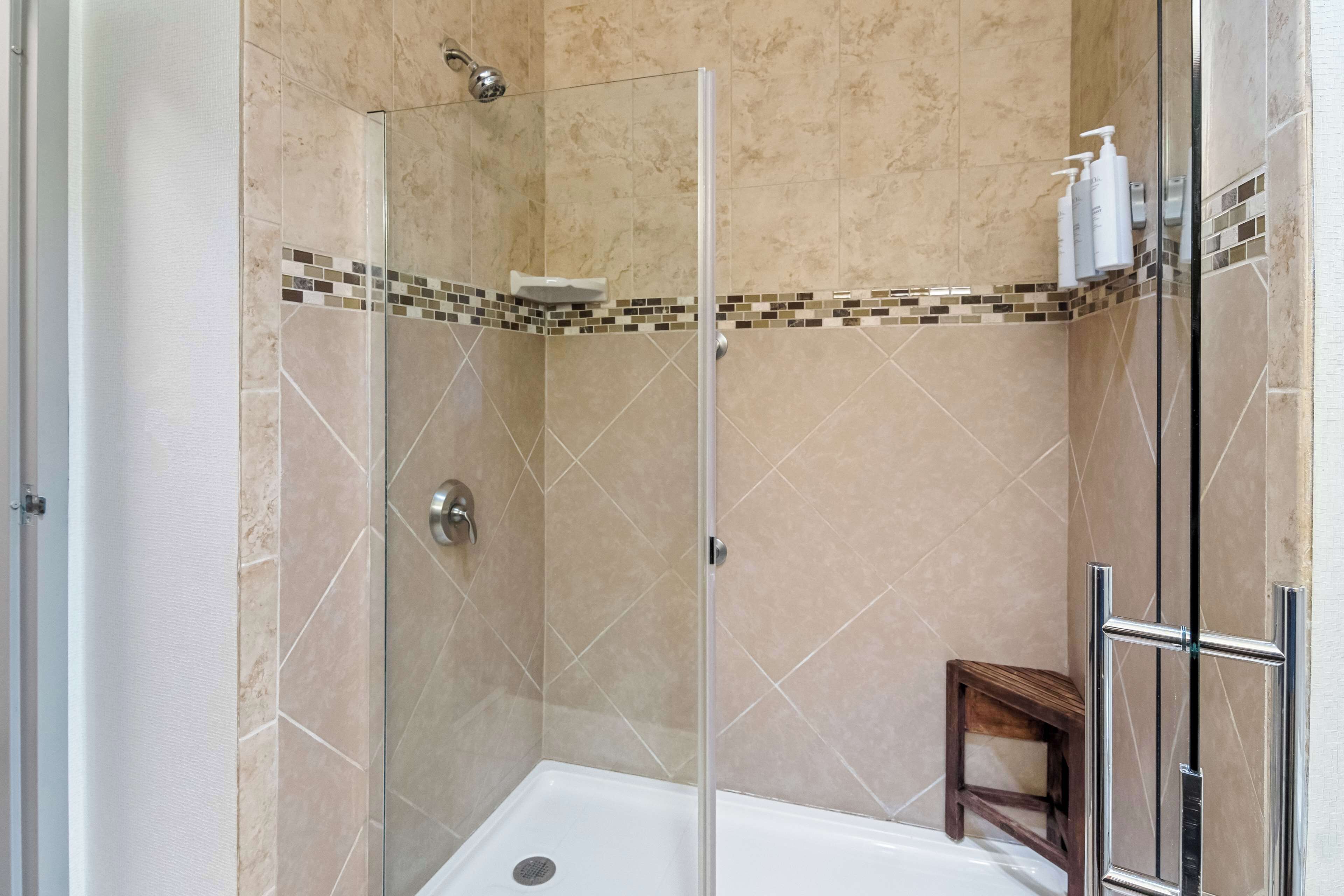 Um chuveiro com divisória de vidro, azulejos bege e um pequeno banquinho de madeira.