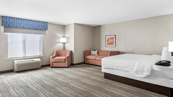 Quarto de hotel com cama, poltrona, sofá, janela com persianas, luminária e radiador.