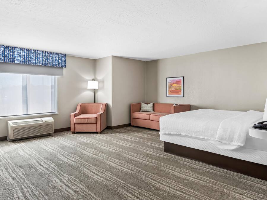 Quarto de hotel com cama, poltrona, sofá, janela com persianas, luminária e radiador.