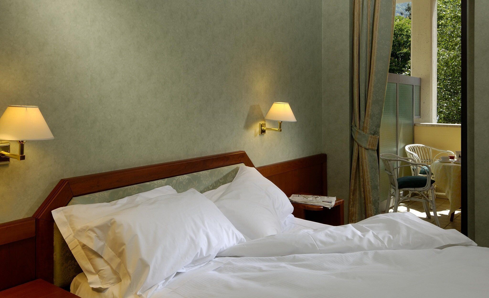 Quarto de hotel com uma cama bem arrumada, duas luminárias fixadas na parede, uma mesinha com uma cadeira e vista para o verde lá fora.