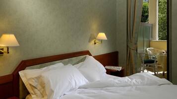 Quarto de hotel com uma cama bem arrumada, duas luminárias fixadas na parede, uma mesinha com uma cadeira e vista para o verde lá fora.