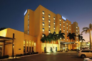 Um hotel com vários andares, com uma placa chamativa da 'City Express', palmeiras e uma área de estacionamento.