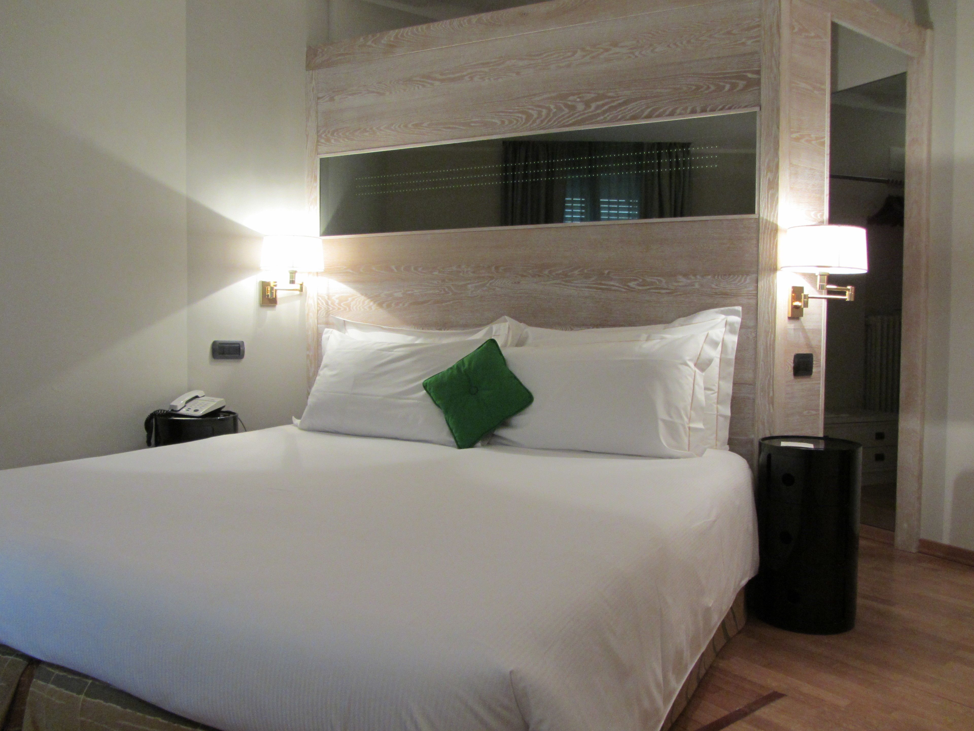 Quarto de hotel com uma cama grande, telefone ao lado da cama, lixeira e luminária fixada na parede.