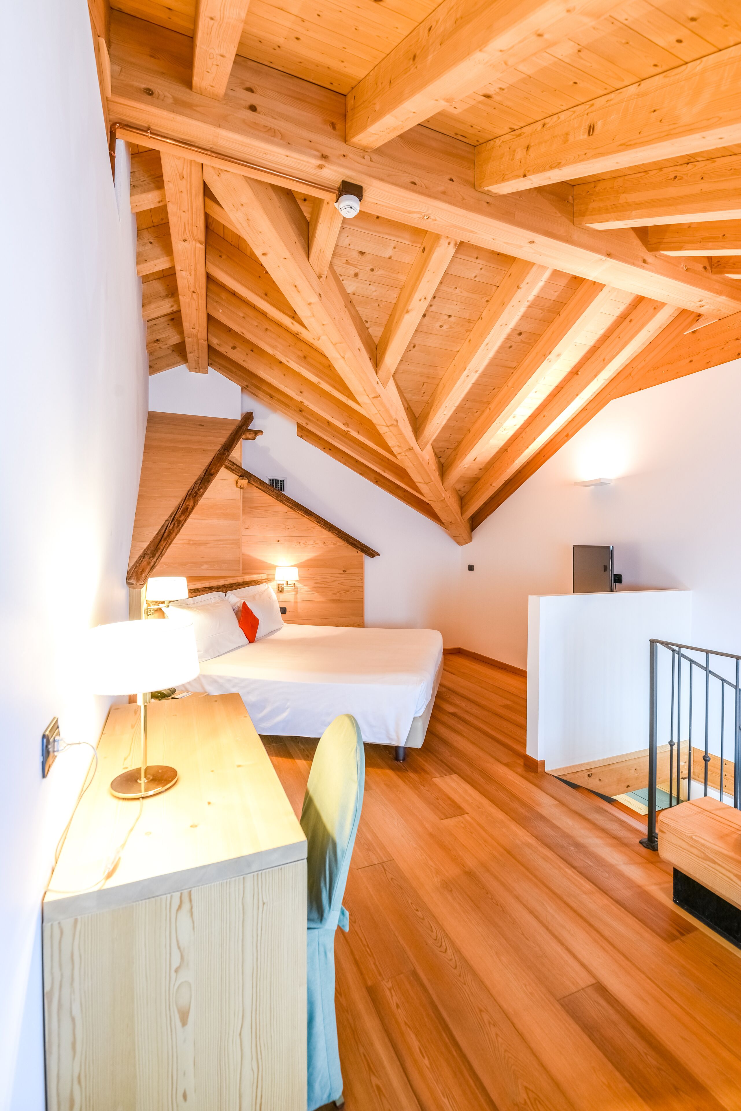 Quarto pequeno e acolhedor, com teto inclinado, vigas de madeira, uma cama, uma escrivaninha e uma cadeira.