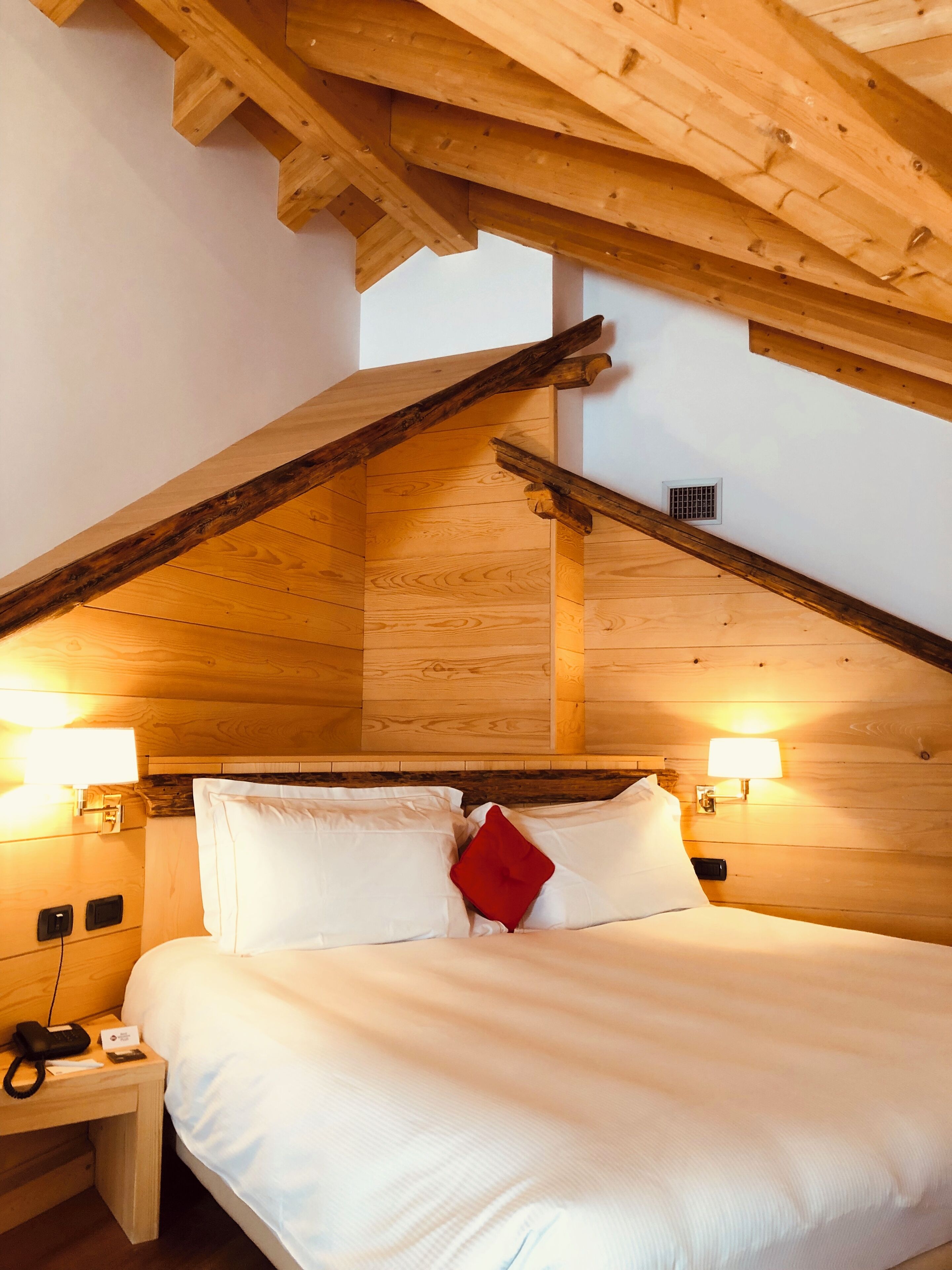 Quarto com teto inclinado, vigas de madeira, cama com roupa de cama branca e travesseiro vermelho, duas luminárias de cabeceira e mesa de cabeceira com telefone e cartão.