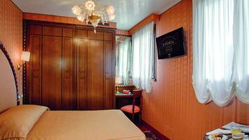 Quarto de hotel com cama, guarda-roupa de madeira, escrivaninha com cadeira, lustre e placa com o nome do hotel.