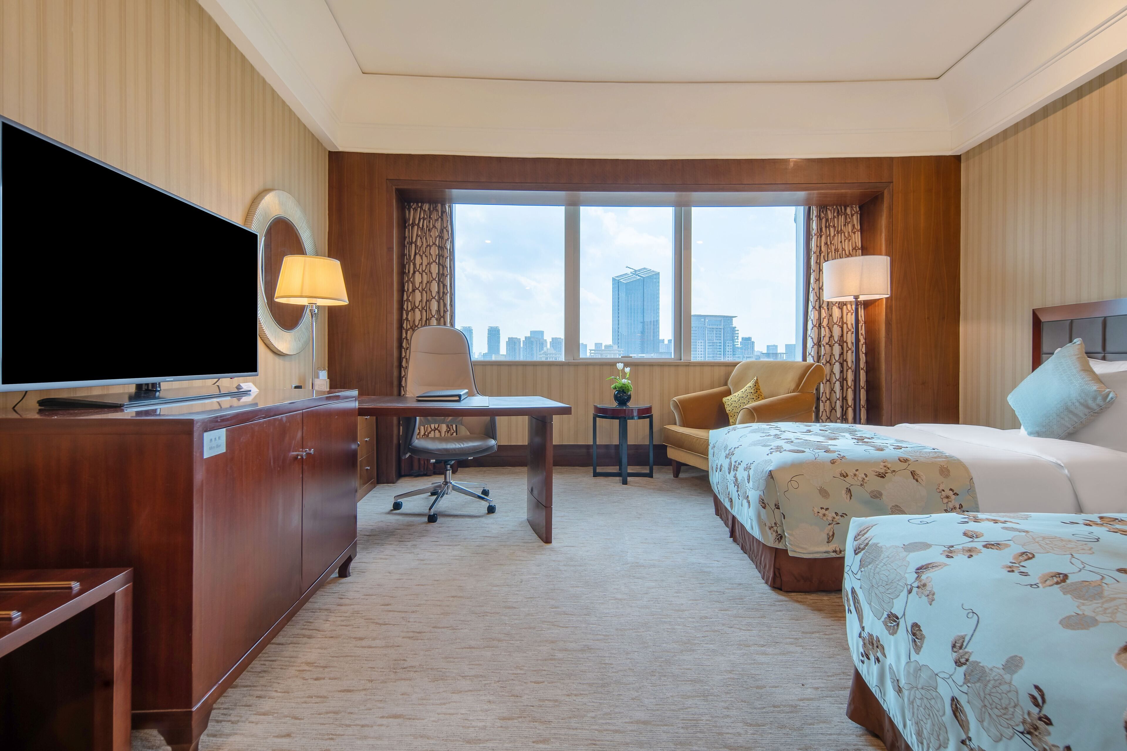 Quarto de hotel com uma grande televisão de tela plana, uma escrivaninha com cadeira, duas camas com colchas florais e vista para o horizonte urbano através de grandes janelas.