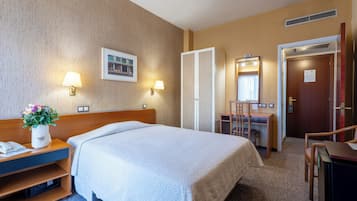 Quarto de hotel com cama, mesa de cabeceira, luminária e escrivaninha com cadeira.