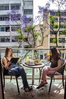 Duas pessoas sentadas à mesa, com um copo de bebida e comida, rodeadas por flores roxas e um edifício ao fundo.