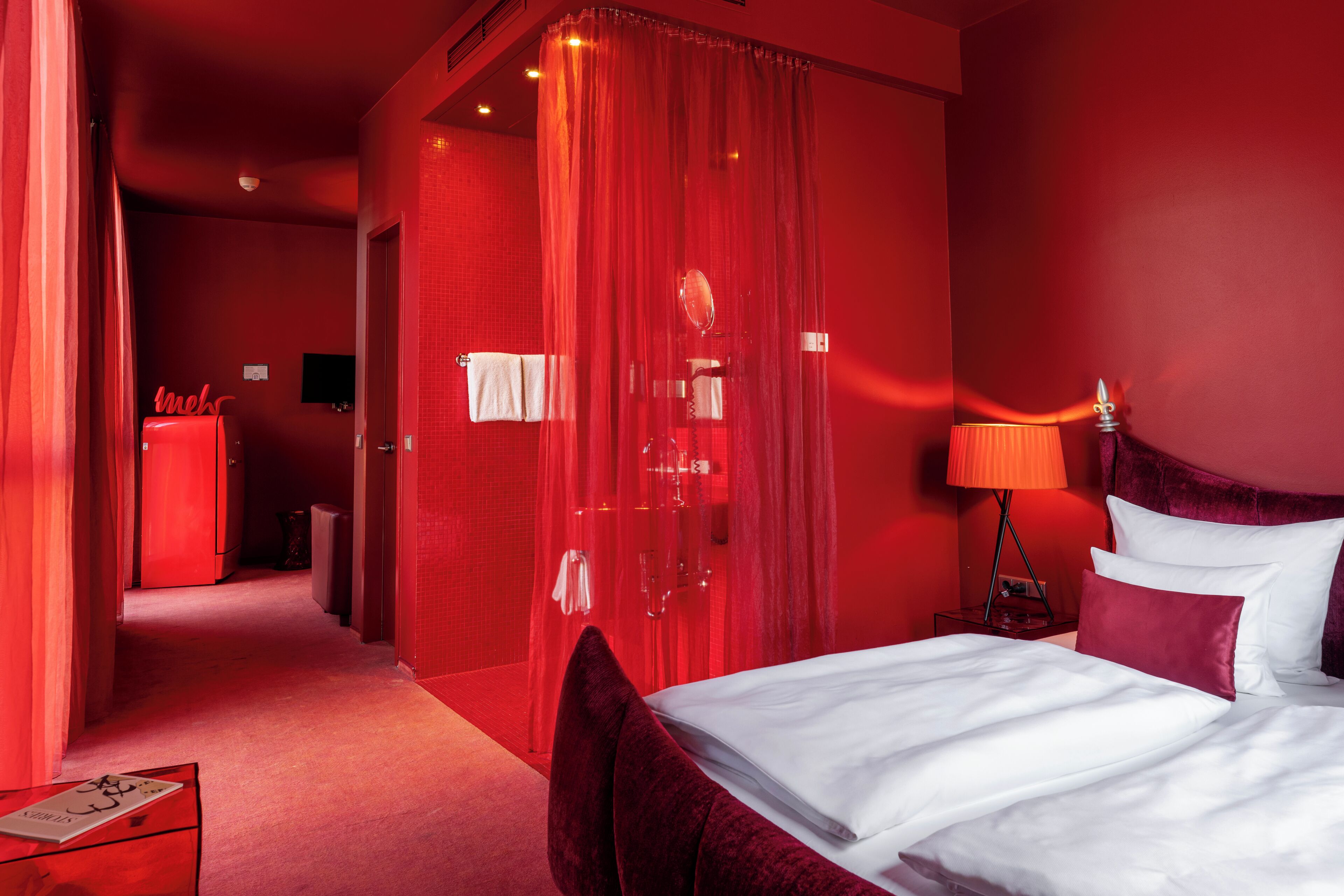 Quarto de hotel em tons de vermelho, com cama, mesa de cabeceira, abajur e espelho.
