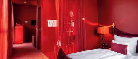 Quarto de hotel em tons de vermelho, com cama, mesa de cabeceira, abajur e espelho.
