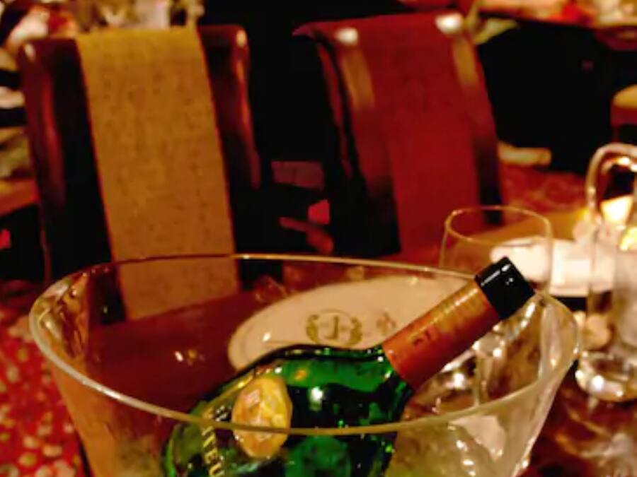 Uma área de bar com um copo contendo uma garrafa de bebida, uma mesa de jantar com cadeiras e uma pessoa ao fundo.