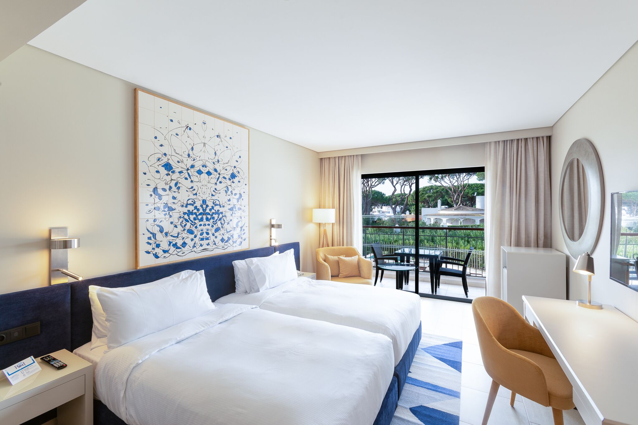 Quarto de hotel com duas camas, uma escrivaninha, uma cadeira, varanda com vista e um quadro abstrato grande na parede.