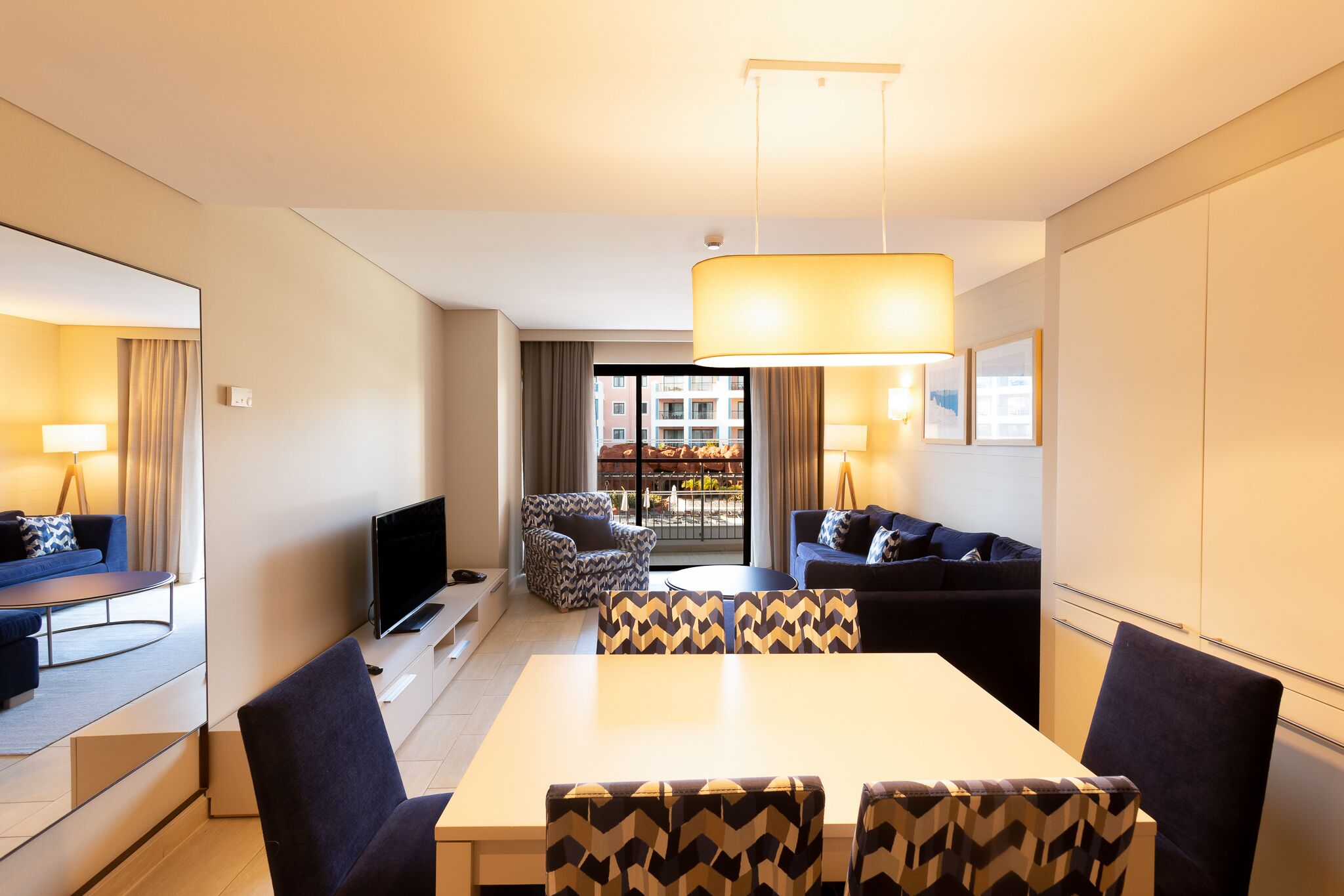 Quarto de hotel moderno com área de refeições, sofá, TV e vista da cidade através de amplas janelas.
