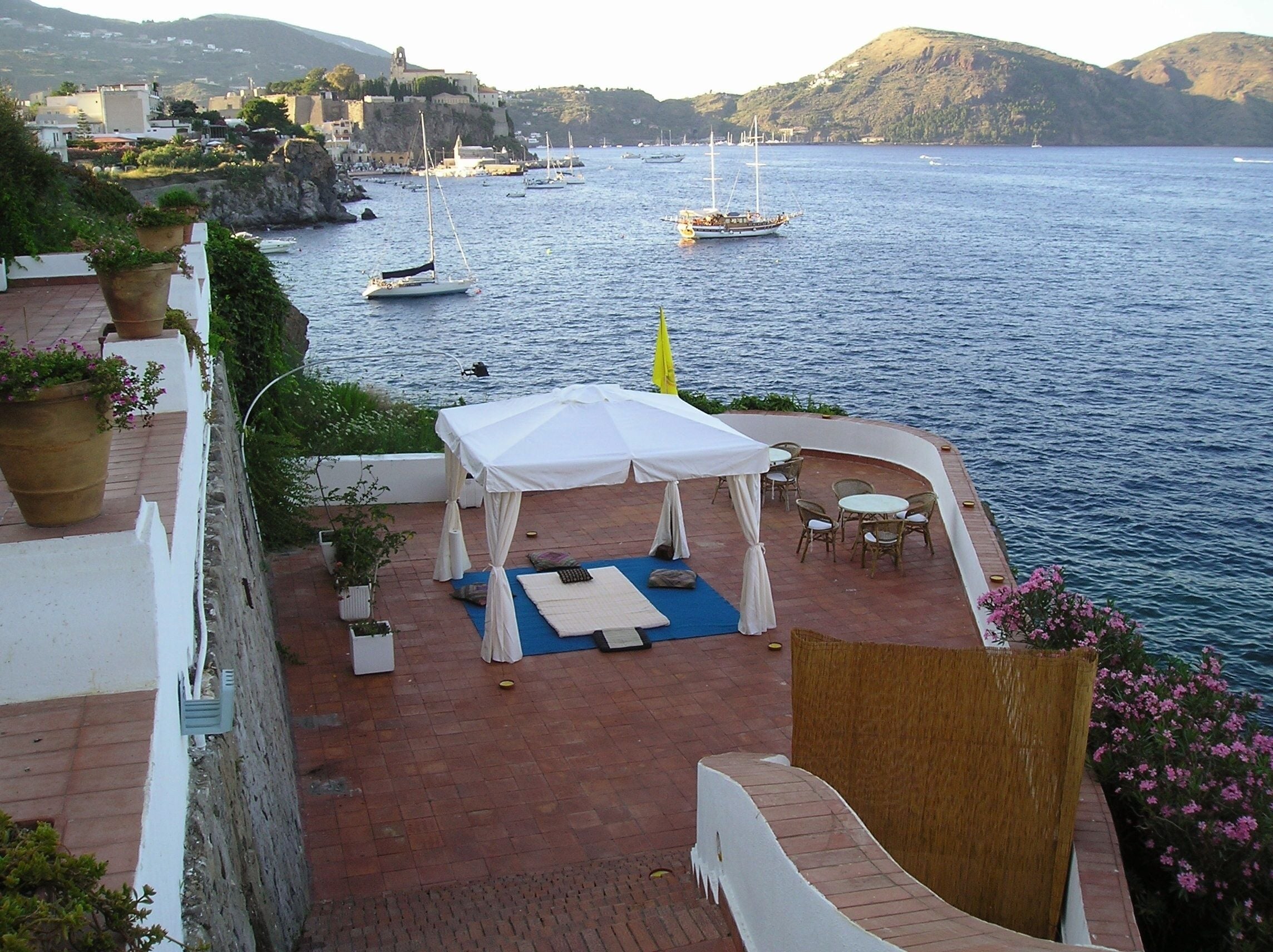 Terraço com toldo branco, mobiliário para área externa e vista para o mar com barcos.
