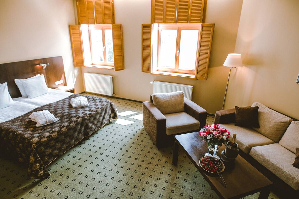 Quarto de hotel com cama, sofá, mesa de centro com flores e duas janelas com persianas.