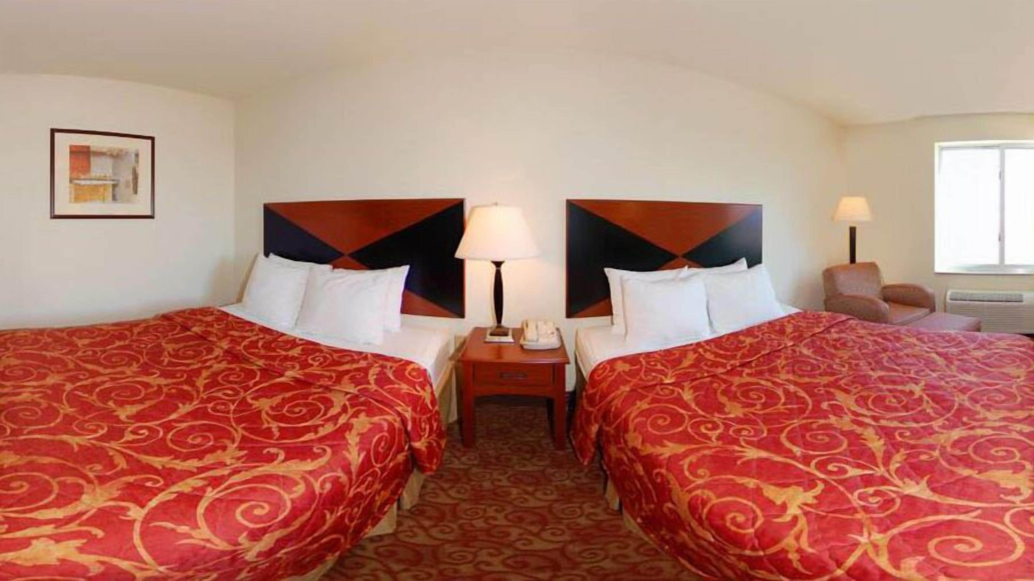 Quarto de hotel com duas camas, cada uma com colcha vermelha estampada, mesa de cabeceira com abajur e uma cadeira.