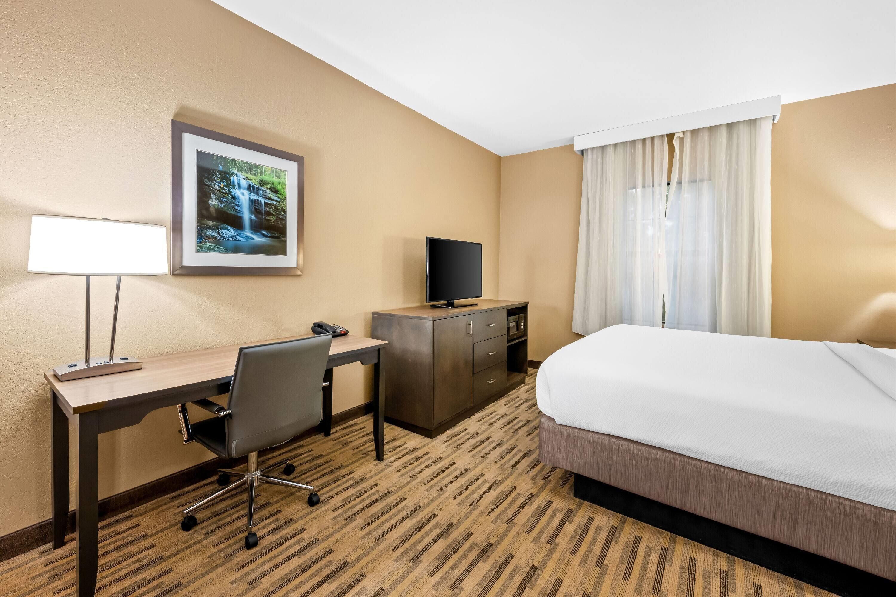 Quarto de hotel com cama, escrivaninha, cadeira, televisão e um quadro na parede.