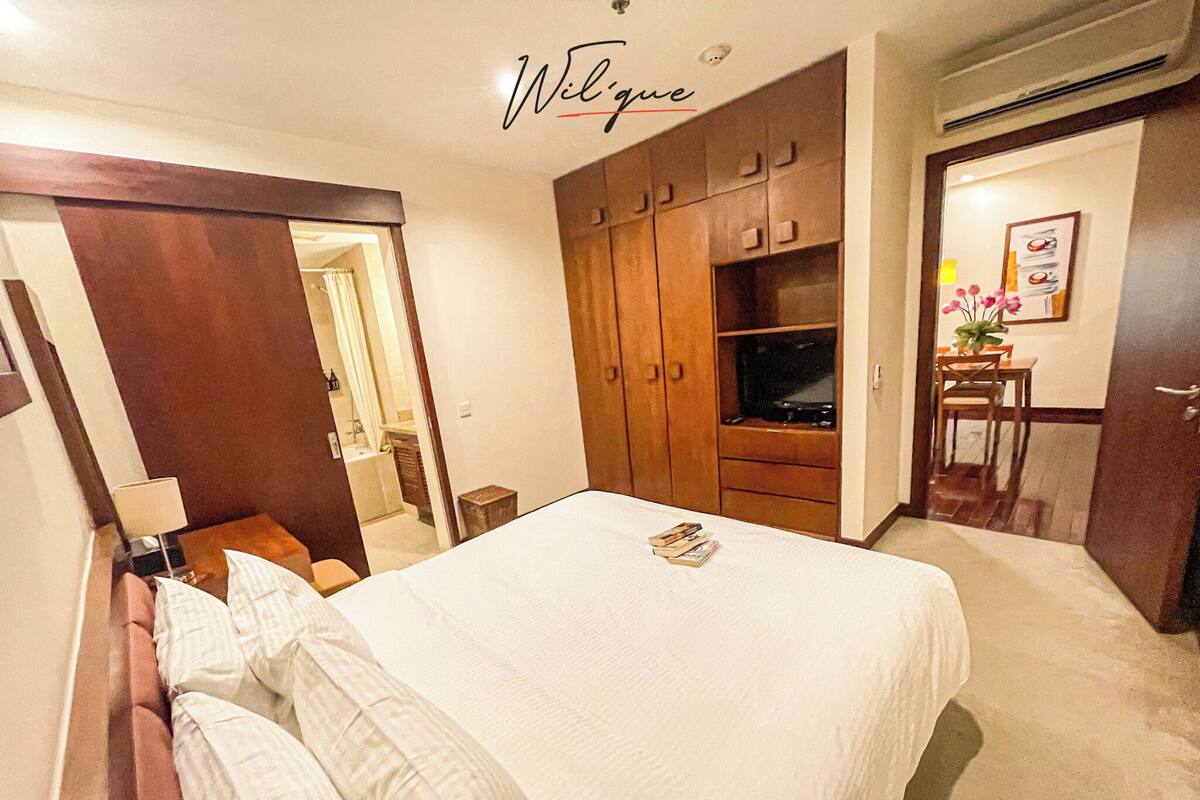 Quarto com cama, guarda-roupa de madeira e uma pequena mesa com luminária.