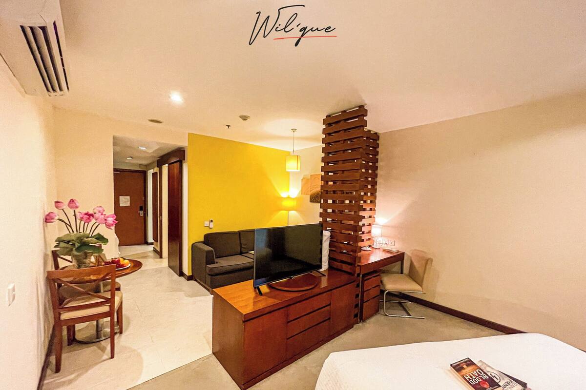 Quarto de hotel com cama, escrivaninha de madeira, cadeira, sofá, televisão e painel decorativo de madeira.