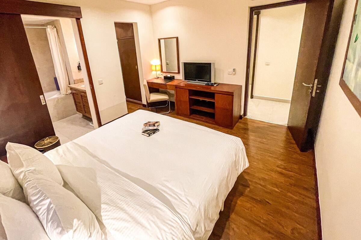 Quarto de hotel com uma cama, uma escrivaninha com televisão, um espelho e uma porta que dá para outro cômodo.