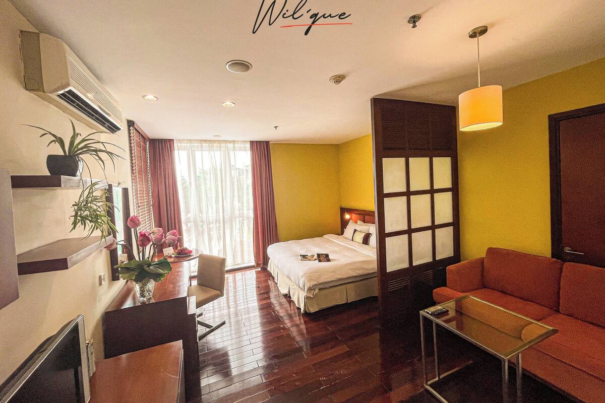 Quarto de hotel com cama, sofá, mesa de jantar com cadeiras, planta em vaso e janela com cortinas.