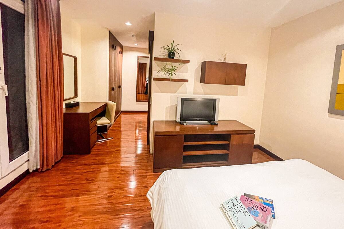 Quarto de hotel com uma cama, uma escrivaninha, uma televisão, uma prateleira com uma planta e uma porta que dá para um corredor.