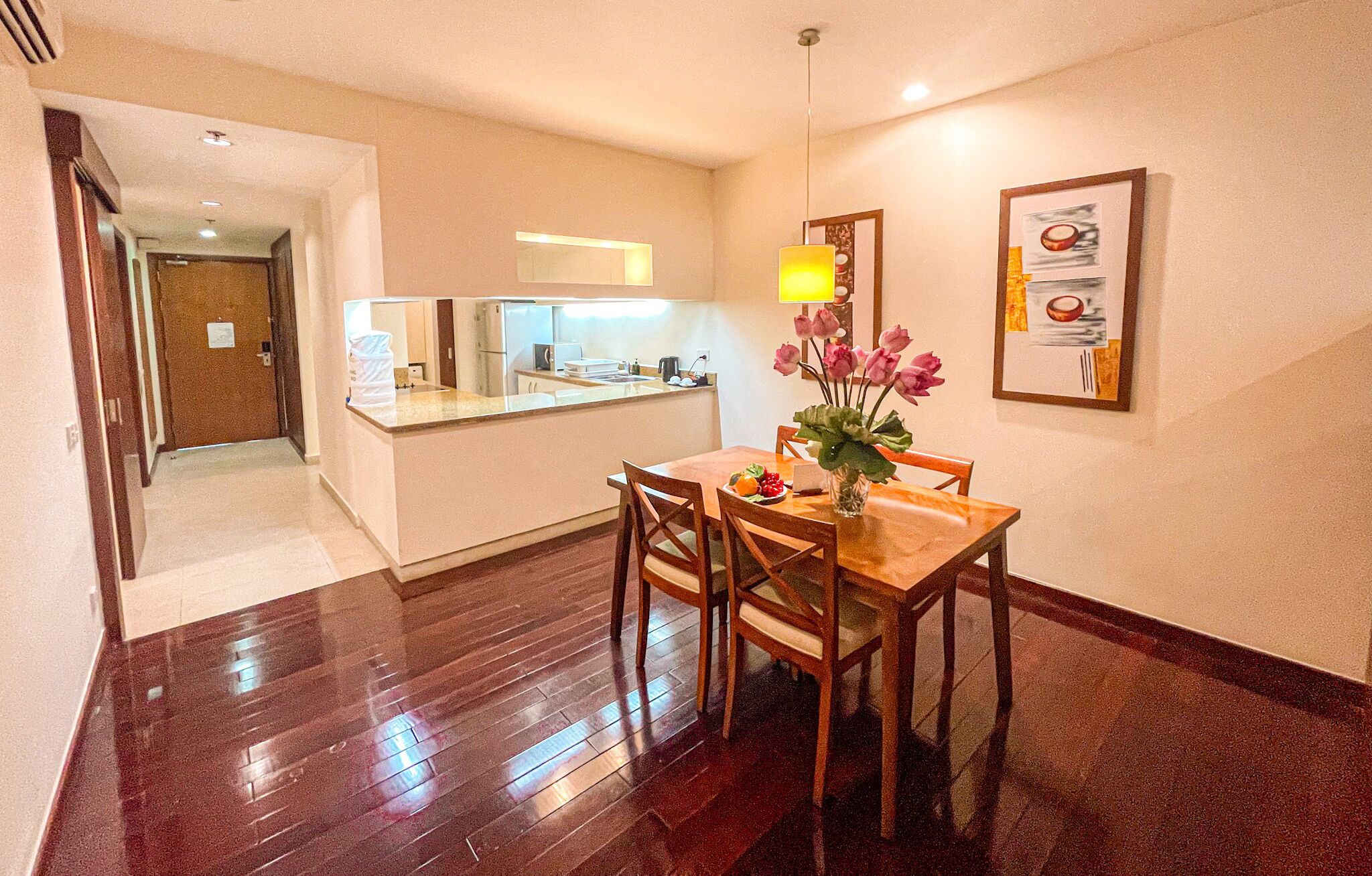 Uma sala de jantar com uma mesa e cadeiras de madeira, um vaso com flores e uma cozinha ao fundo.
