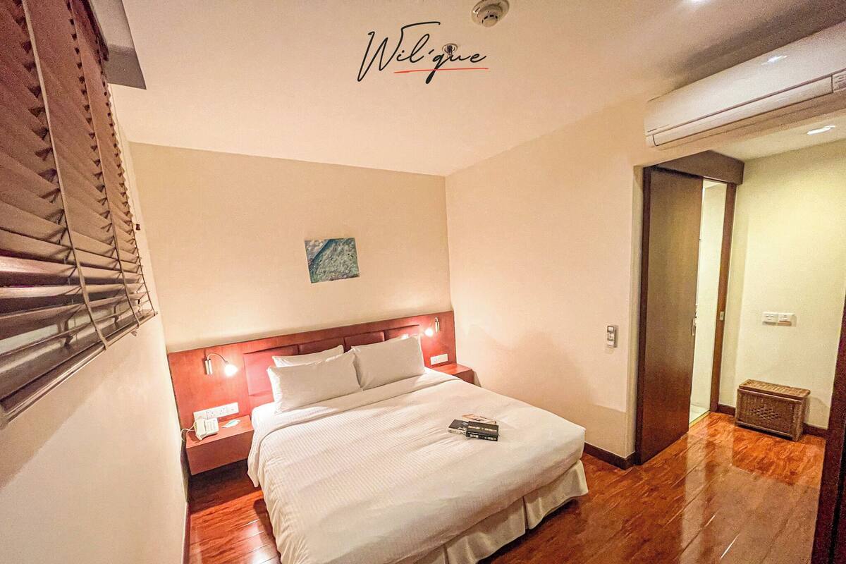 Quarto de hotel com piso de madeira, cama com lençóis brancos, mesa de cabeceira com abajur e um quadro na parede.