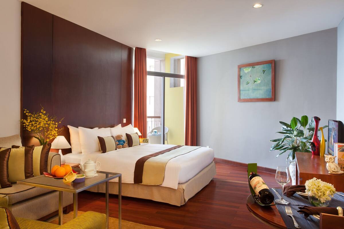 Quarto de hotel com cama, mesa de cabeceira, luminária, flores e uma garrafa de vinho.
