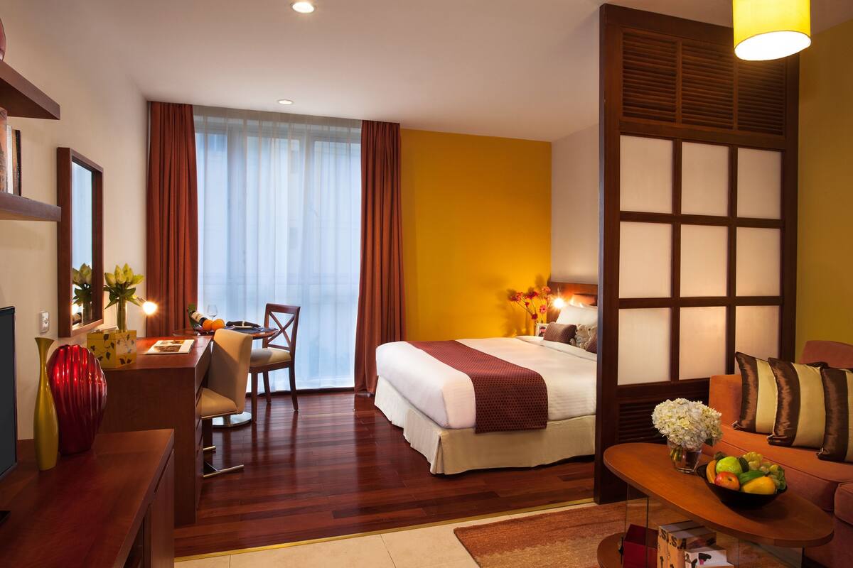 Quarto de hotel com cama, secretária com cadeira, sofá, mesa de jantar e janela com cortinas.