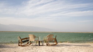 Uma praia com duas cadeiras de vime e uma mesa, com vista para o mar tranquilo e montanhas ao longe.