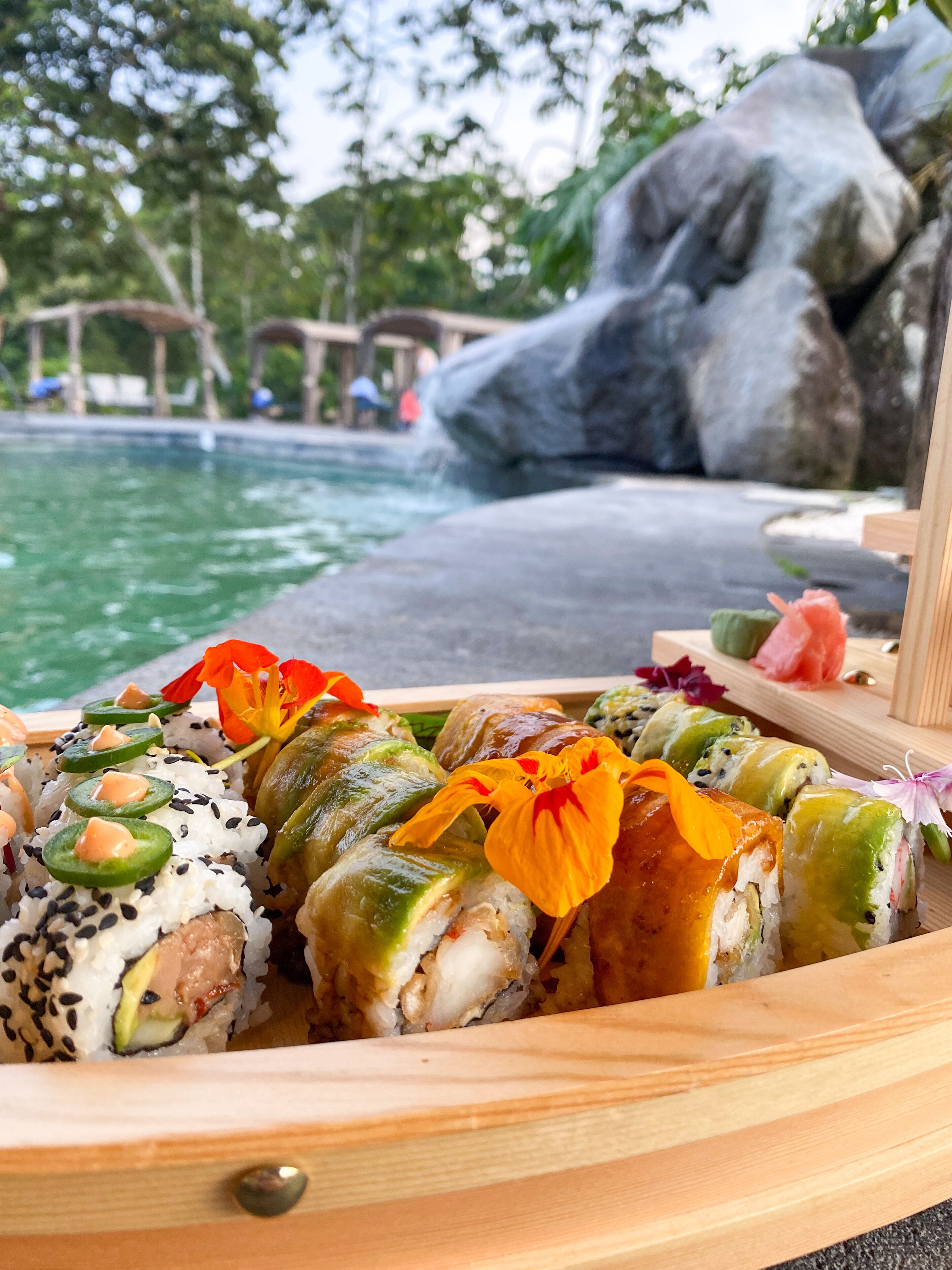 Uma bandeja com rolinhos de sushi e flores decorativas, colocada sobre uma superfície de madeira, perto de uma piscina com pedras e vegetação ao fundo.
