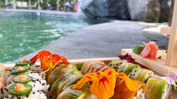 Uma bandeja com rolinhos de sushi e flores decorativas, colocada sobre uma superfície de madeira, perto de uma piscina com pedras e vegetação ao fundo.