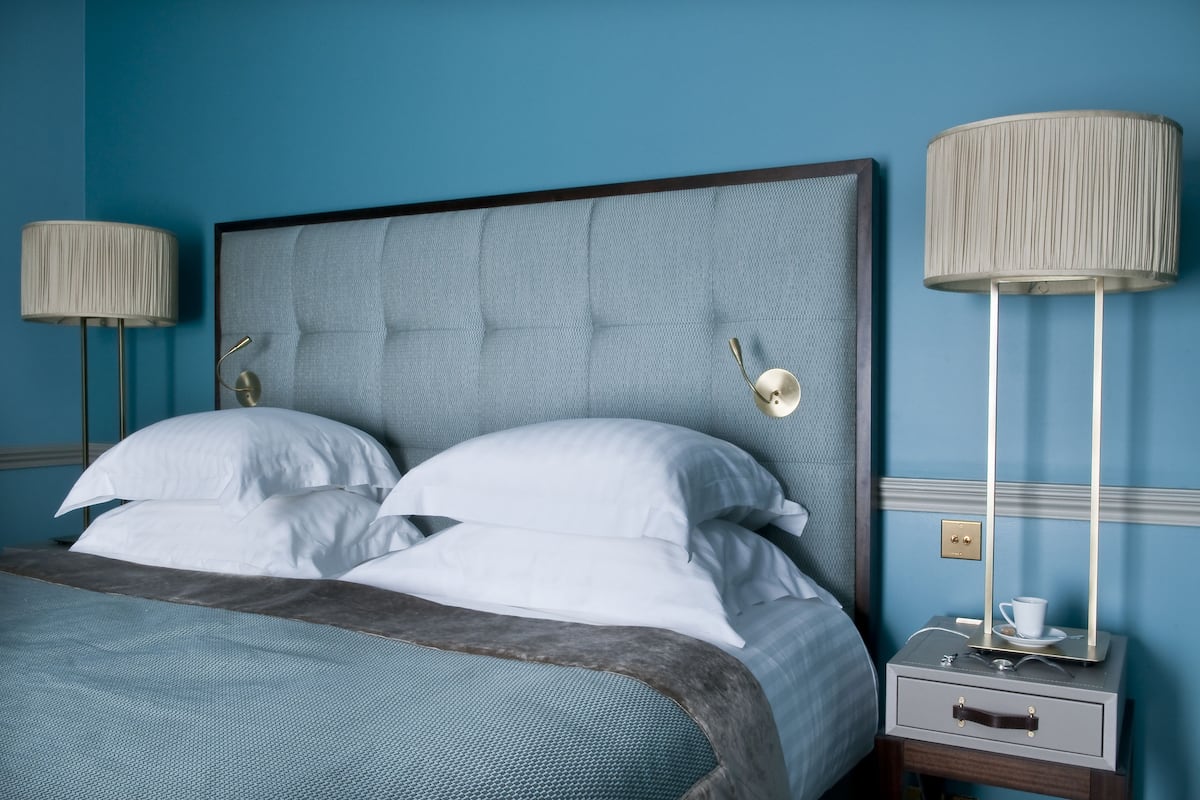 Uma cama bem arrumada com lençóis brancos, cabeceira com design acolchoado, duas luminárias de cabeceira e uma mesa de cabeceira com xícara e pires.