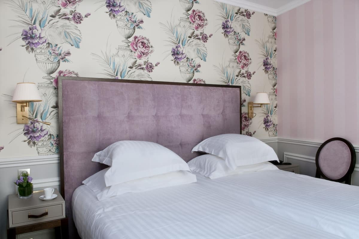 Uma cama bem arrumada com lençóis brancos, cabeceira estofada em roxo e papel de parede floral.