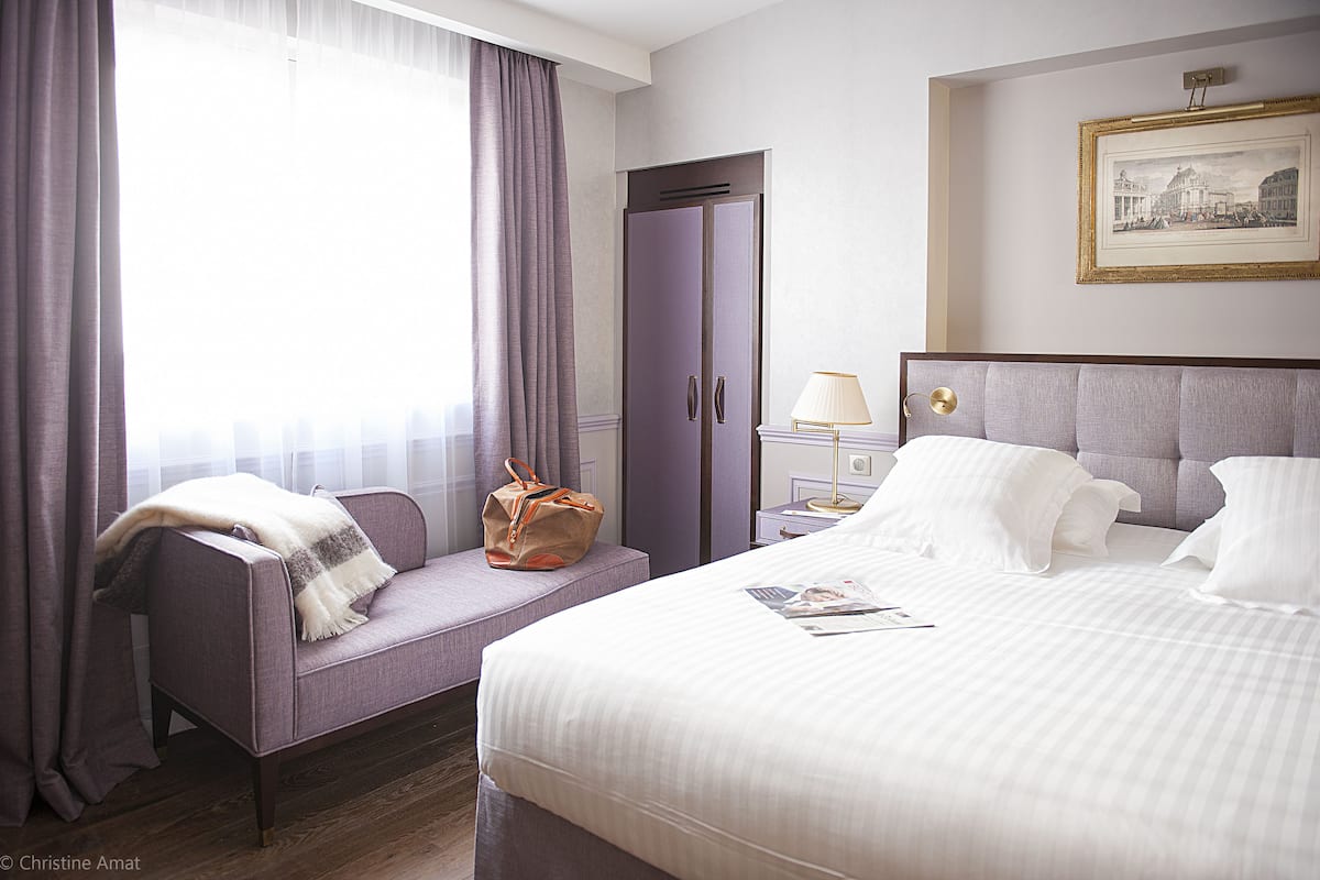 Quarto de hotel com uma cama, um sofá, uma mesa de cabeceira, um abajur e uma porta.