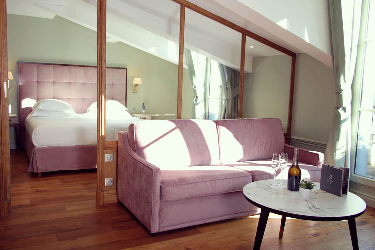 Quarto de hotel com cama, sofá, uma mesinha com uma garrafa e copos, e um espelho refletindo o ambiente.