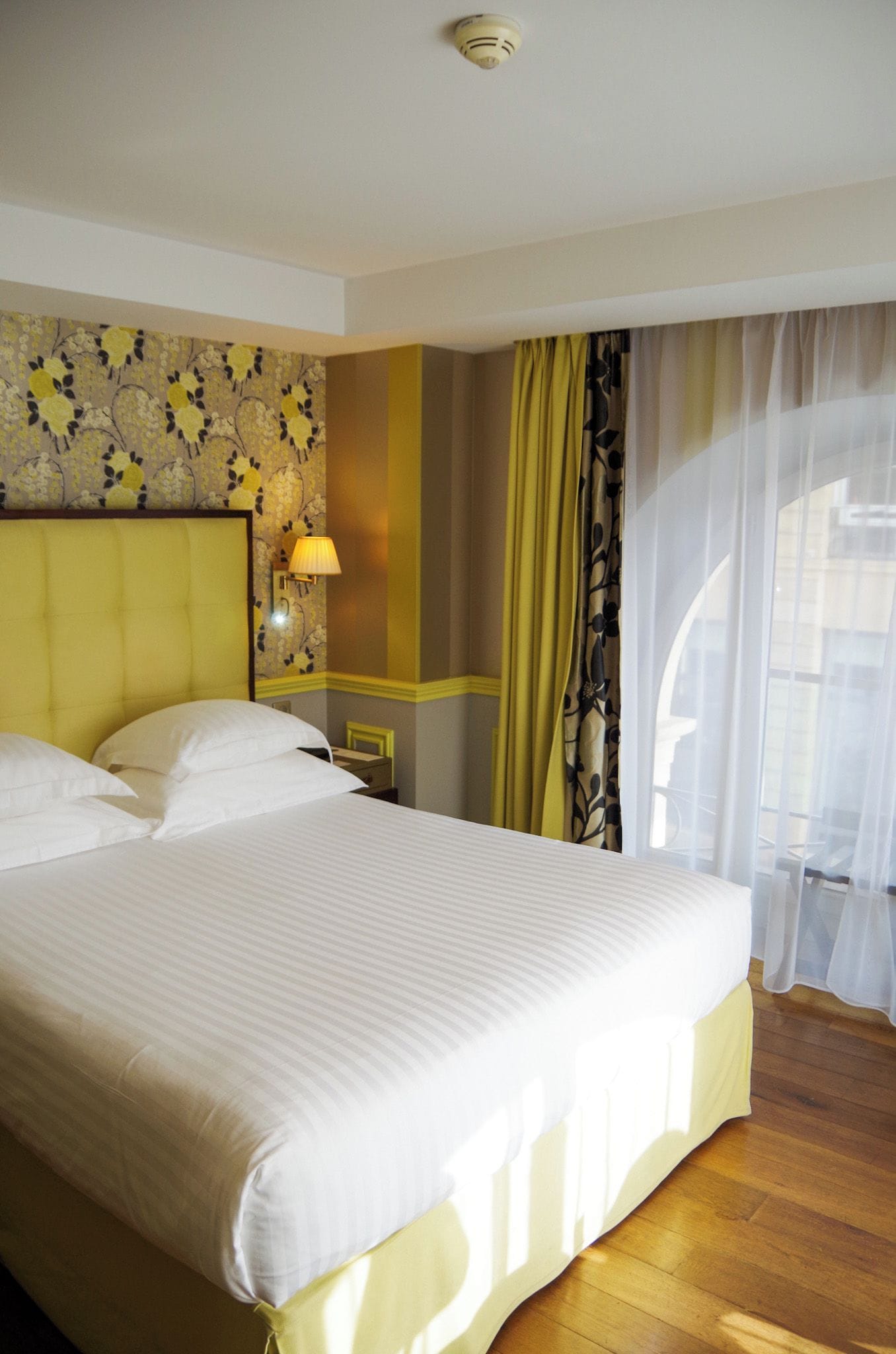 Quarto de hotel com uma cama grande, cabeceira, mesa de cabeceira, abajur e janela com cortinas.
