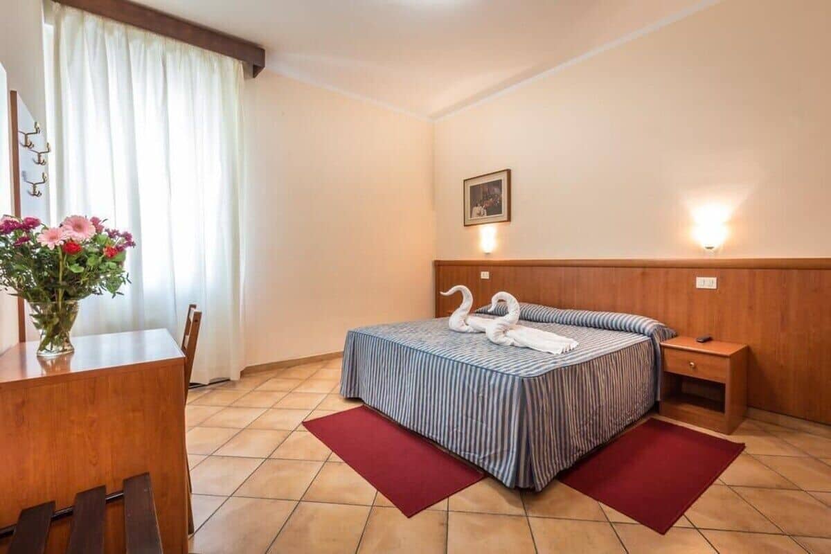 Quarto de hotel com cama, escrivaninha, cadeira, um vaso de flores e um quadro na parede.