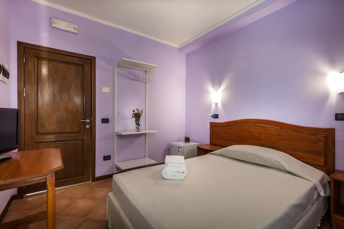 Quarto com porta de madeira, uma cama com cabeceira de madeira, uma mesinha de cabeceira pequena, um criado-mudo e uma luminária fixada na parede, além de uma prateleira com um vaso.