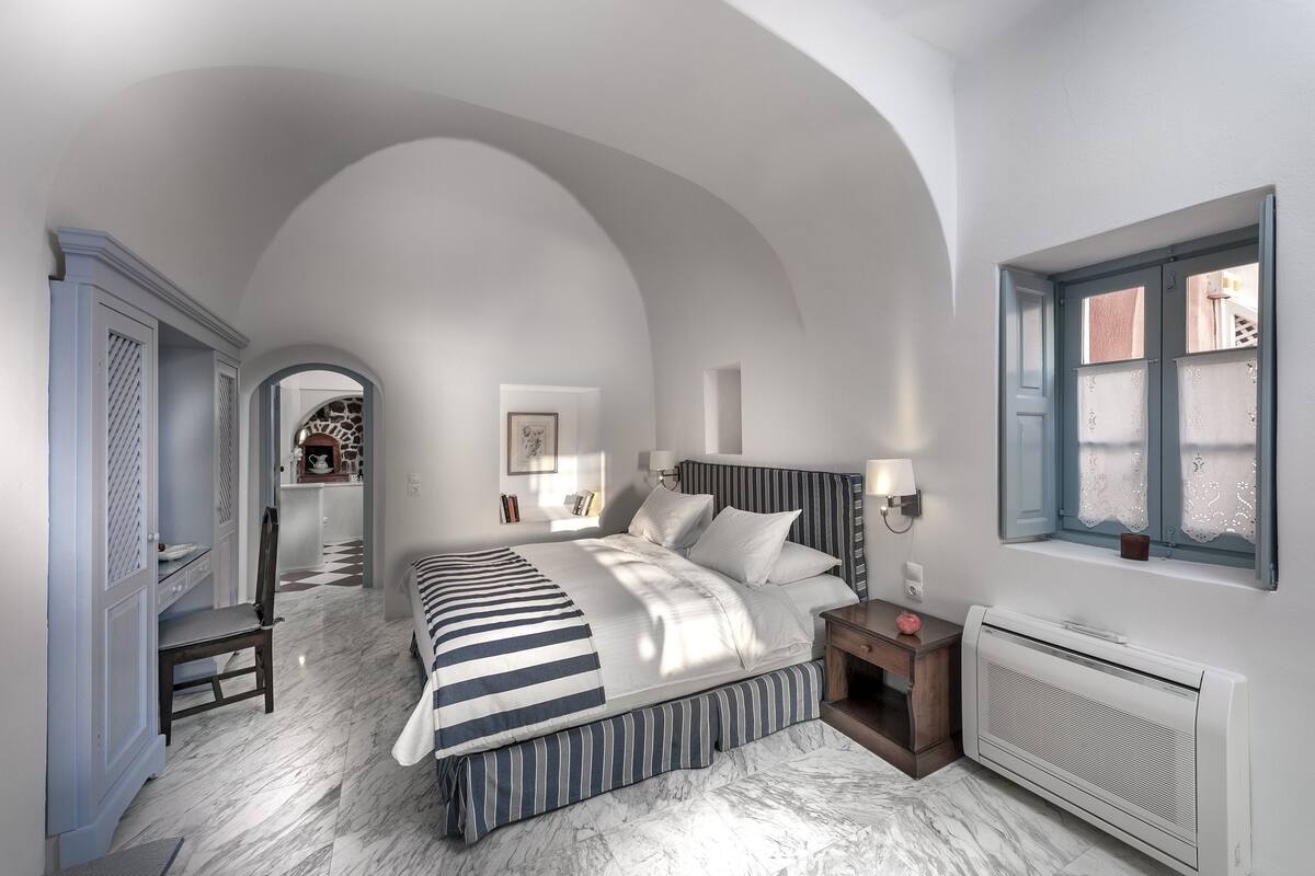 Quarto com cama, mesa de cabeceira, radiador e janela com cortinas.