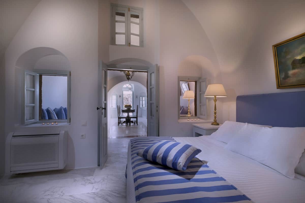 Quarto de hotel com cama, mesa de cabeceira, abajur, pintura e uma porta arqueada que dá para uma sala de jantar.