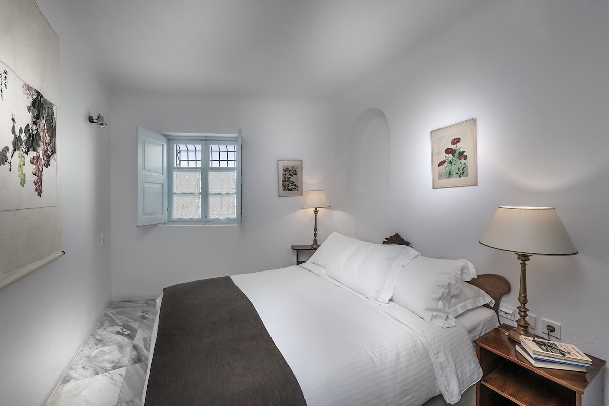 Cama bem arrumada com lençóis brancos e manta escura, mesa de cabeceira com abajur, e pequeno criado-mudo com livros e abajur.
