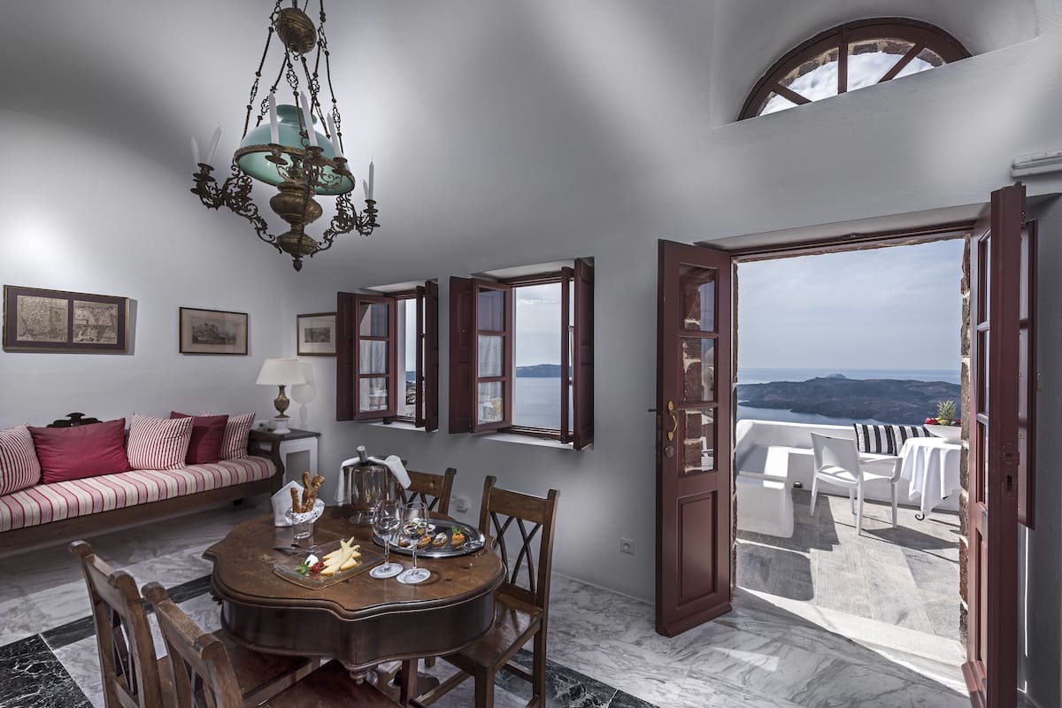 Uma área de refeições com uma mesa redonda, cadeiras e um candelabro. Há vista para o mar pelas janelas abertas.