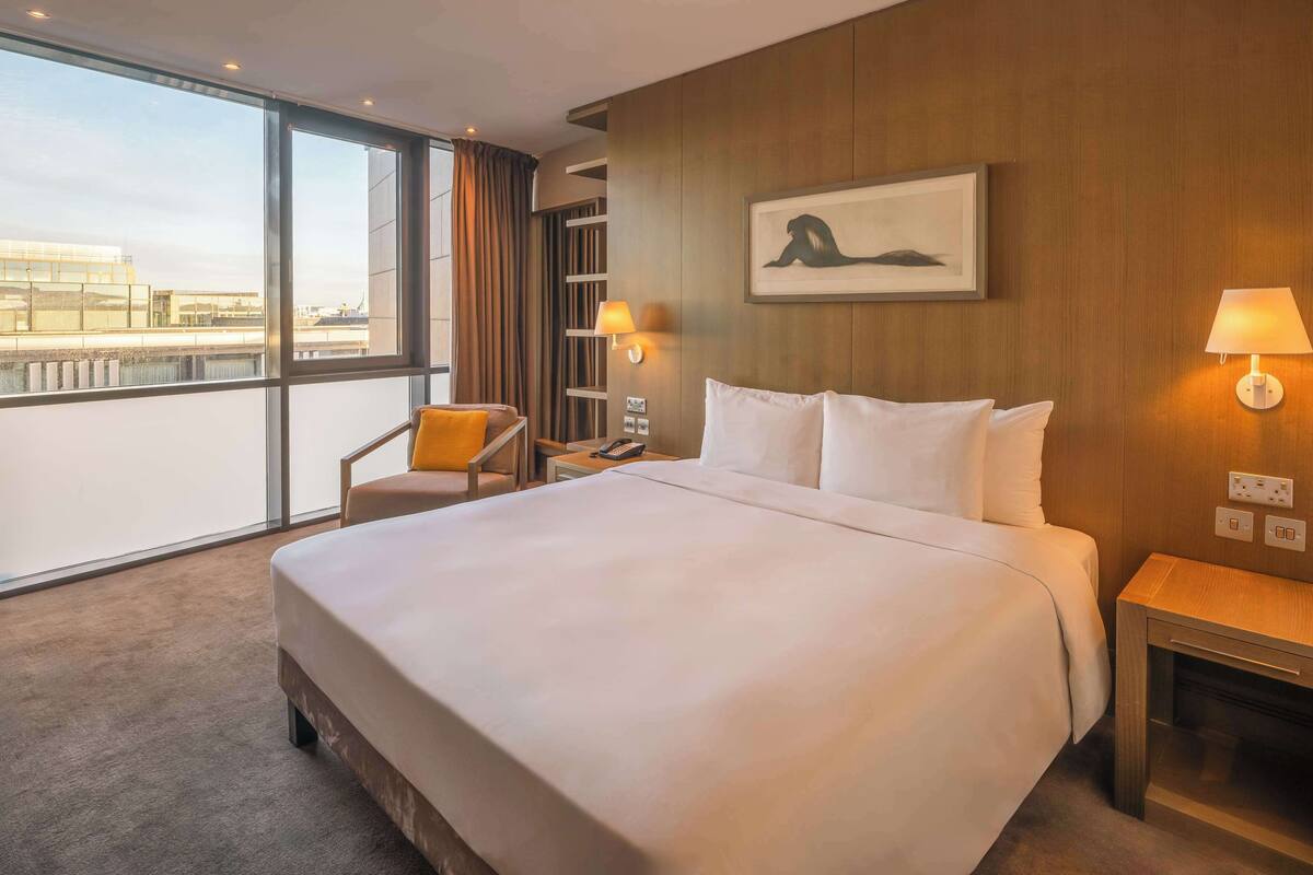 Quarto de hotel com uma cama grande, uma cadeira, uma mesinha de cabeceira, um abajur e um quadro na parede.