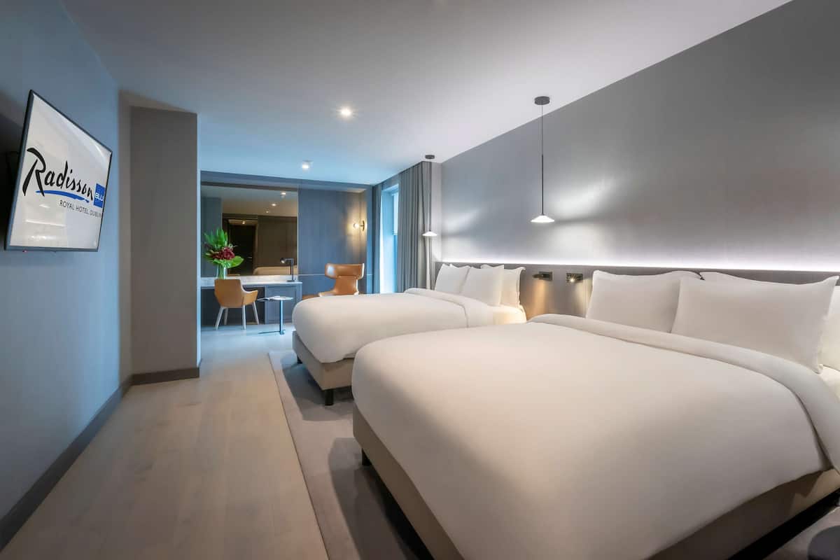 Quarto de hotel moderno com duas camas, área de estar com uma cadeira e mesa, e vista interna para o quarto.