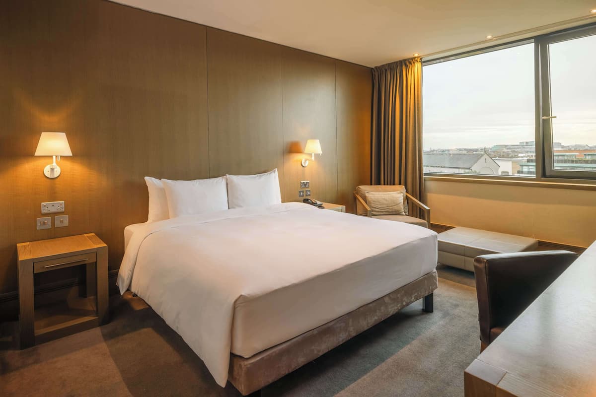 Quarto de hotel com uma cama grande, duas luminárias de cabeceira, uma escrivaninha com cadeira e vista para a cidade pela janela.