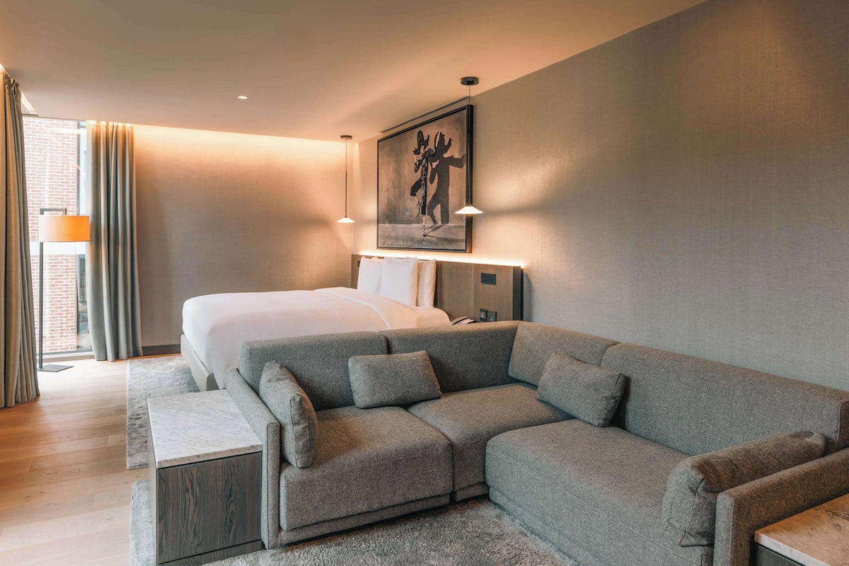 Quarto de hotel moderno com uma cama grande, um sofá cinzento, uma mesinha e um quadro na parede.