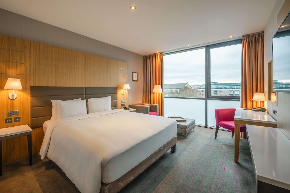 Quarto de hotel com uma cama grande, duas mesas de cabeceira com abajures, uma escrivaninha com cadeira e uma janela ampla com cortinas.